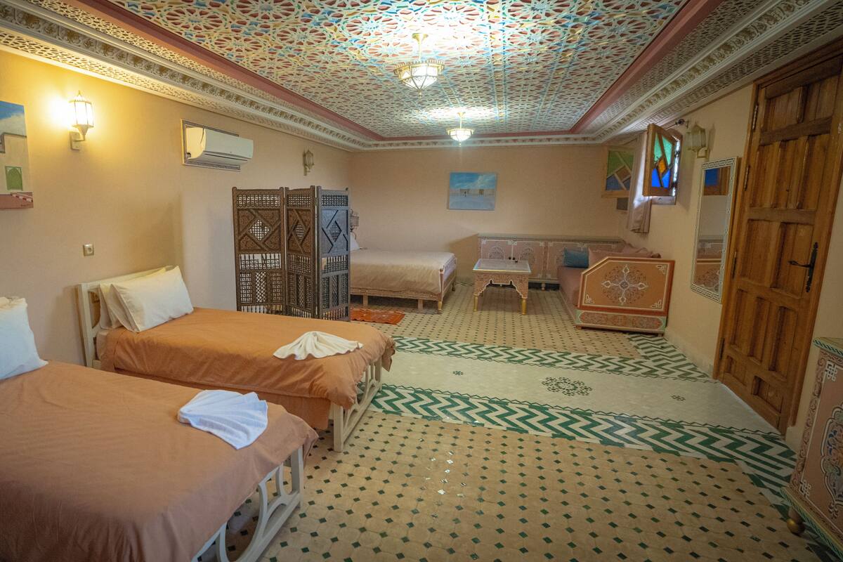 Quarto de hotel com duas camas, um sofá, uma mesinha e teto decorativo.