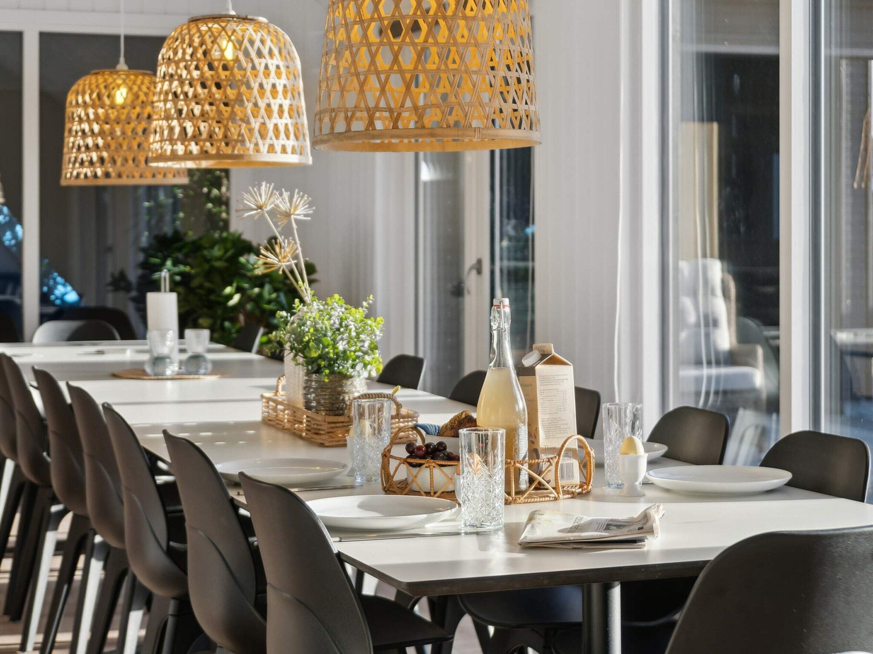 Uma sala de jantar com uma mesa comprida posta para refeição, com cestos de vime, copos e uma garrafa de vinho.