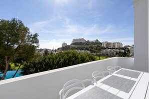 Uma varanda com cadeiras e mesa brancas, com vista para uma piscina e edifícios ao longe.