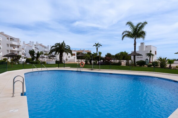 Piscina com água azul cristalina, rodeada por um prédio branco com varandas, palmeiras e céu claro.