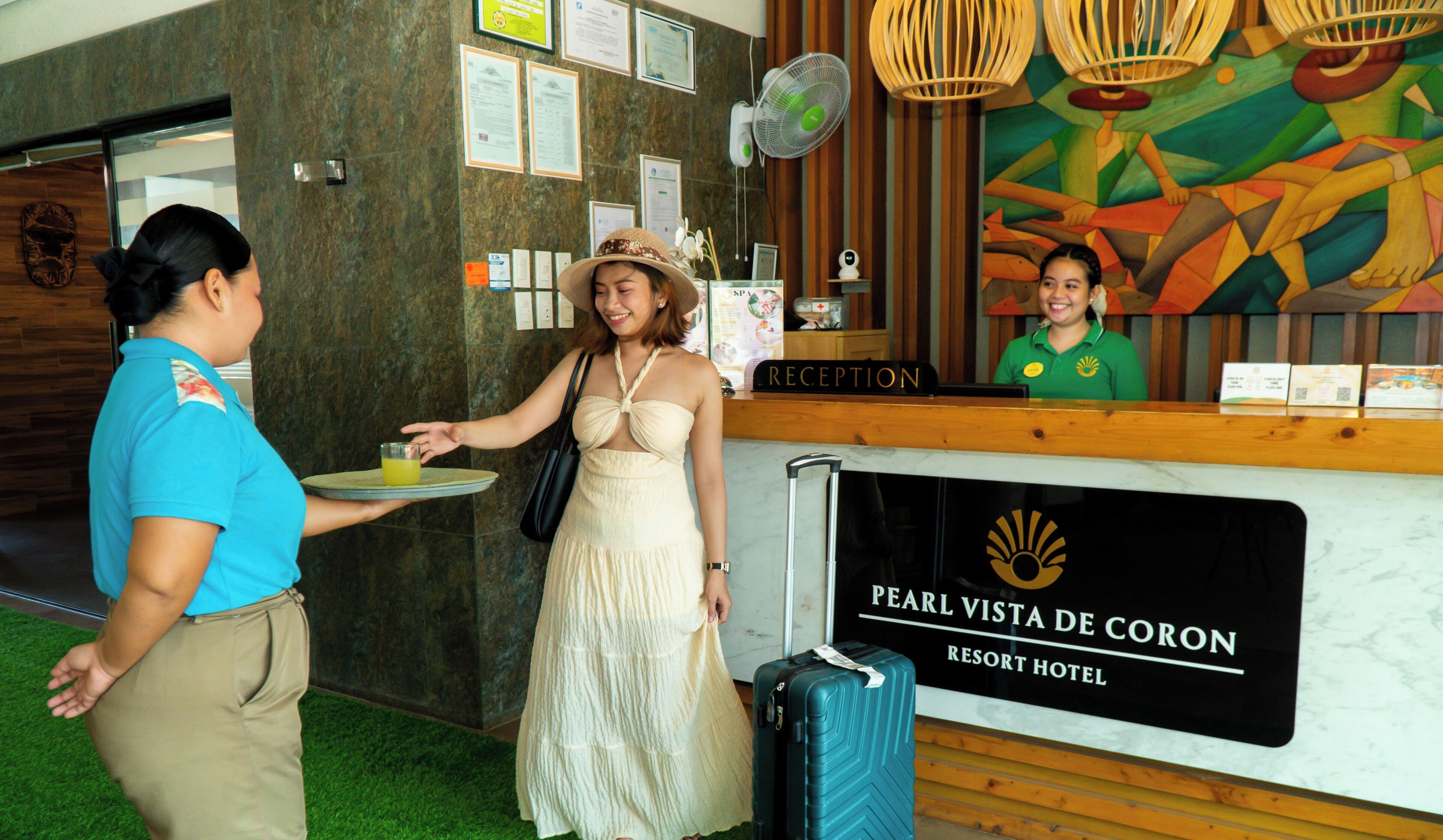 Bancada de recepção no Pearl Vista De Coron Resort Hotel, com uma placa indicando o nome e uma área de recepção com ventilador e obras de arte na parede.