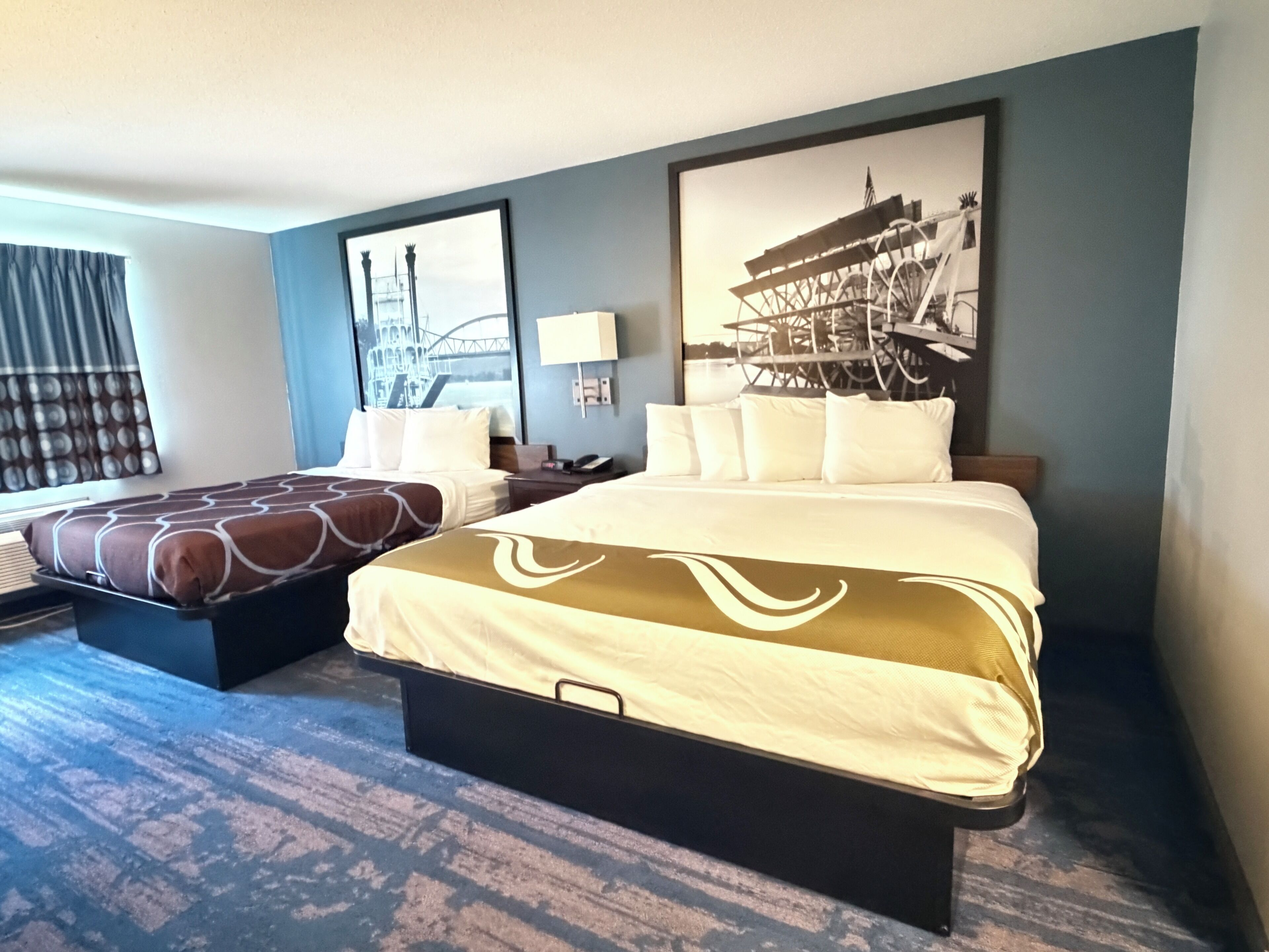 Quarto de hotel com duas camas, uma janela grande com cortinas, uma luminária fixada na parede e um quadro emoldurado.