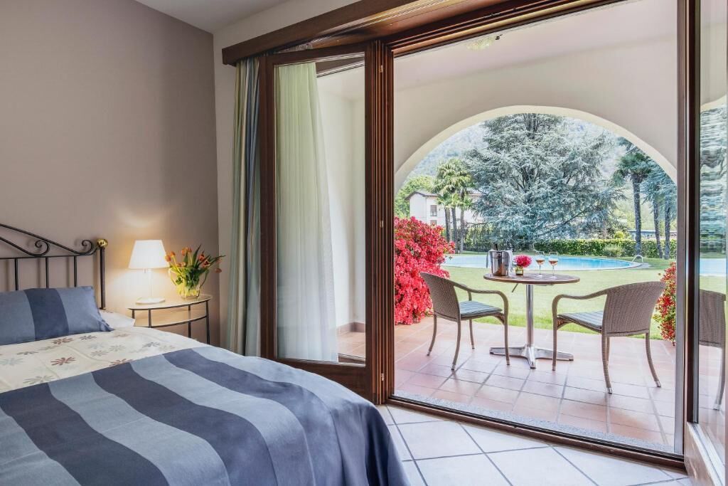 Quarto de hotel com cama, mesa de cabeceira, luminária e vista para um pátio externo com mesa, cadeiras e piscina.