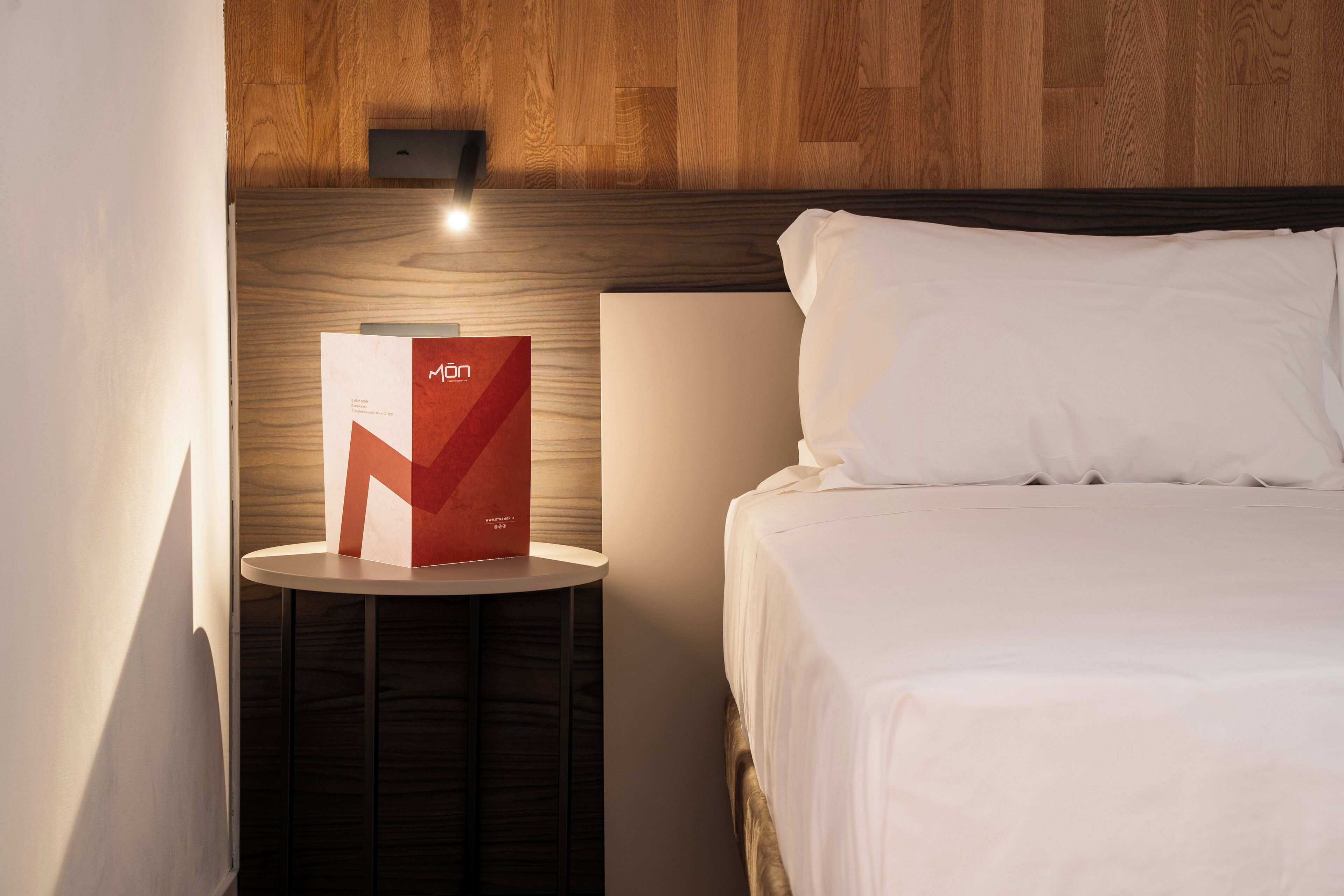 Cama bem arrumada com lençóis brancos, mesa de cabeceira redonda com um livro vermelho, e cabeceira de madeira.