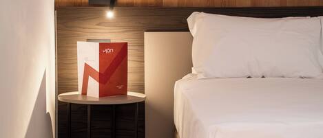 Cama bem arrumada com lençóis brancos, mesa de cabeceira redonda com um livro vermelho, e cabeceira de madeira.