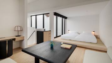 Quarto moderno e minimalista com piso de madeira, uma mesa preta com uma planta, um sofá branco com almofadas e uma escrivaninha com abajur.