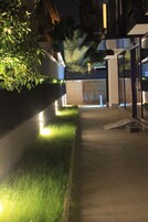 Área externa iluminada à noite, com caminho, grama e um edifício com portas de vidro.