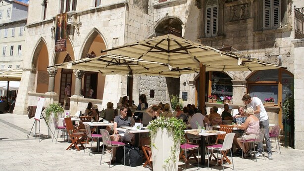 Um café ao ar livre com mesas e cadeiras, com vista para um edifício histórico de pedra com arcos e varanda.
