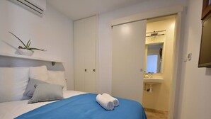 Quarto com cama, mesa de cabeceira, planta, guarda-roupa e banheiro visível por uma porta aberta.