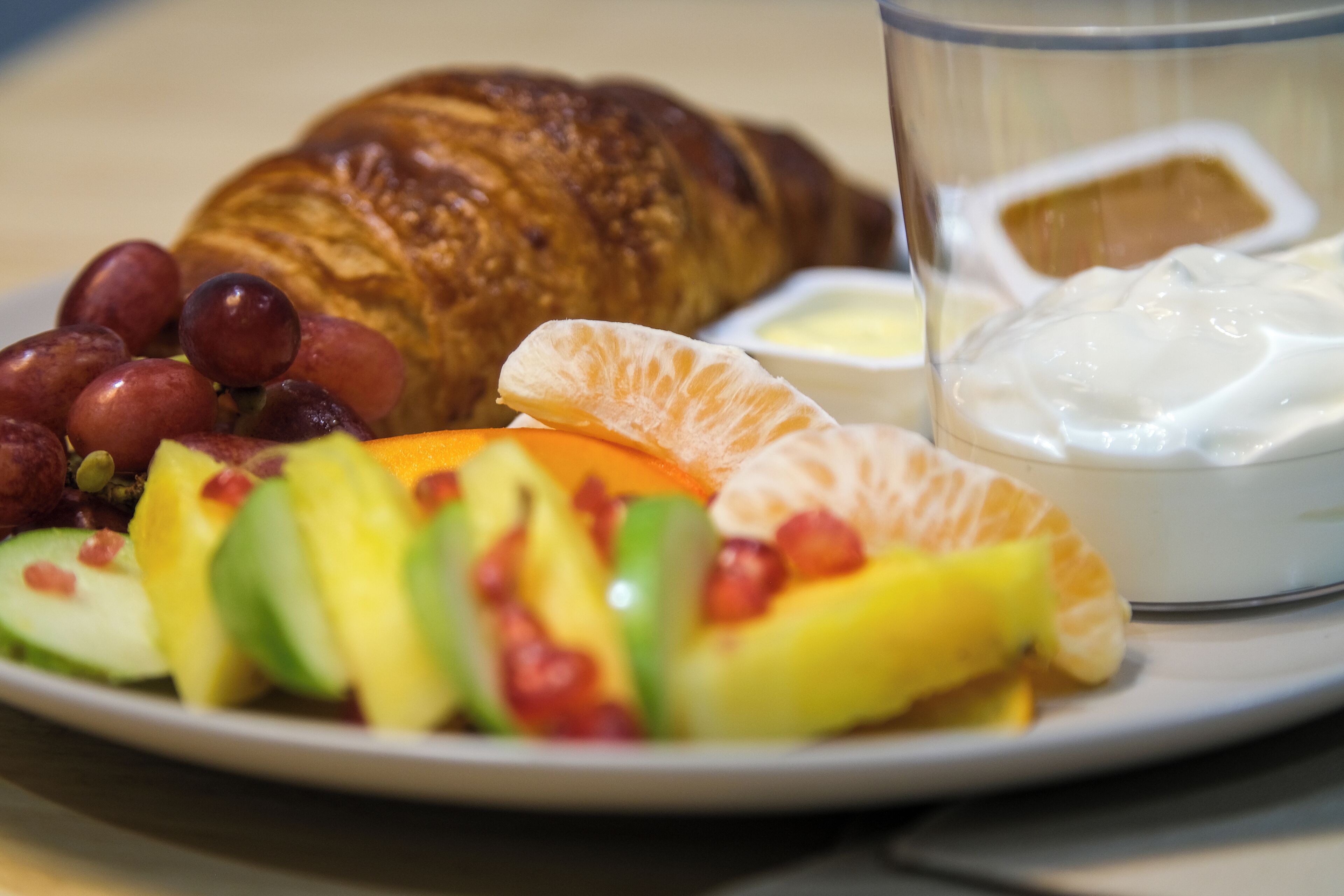Prato com um croissant, uvas, frutas fatiadas e um copo de leite com uma colher.