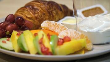 Prato com um croissant, uvas, frutas fatiadas e um copo de leite com uma colher.