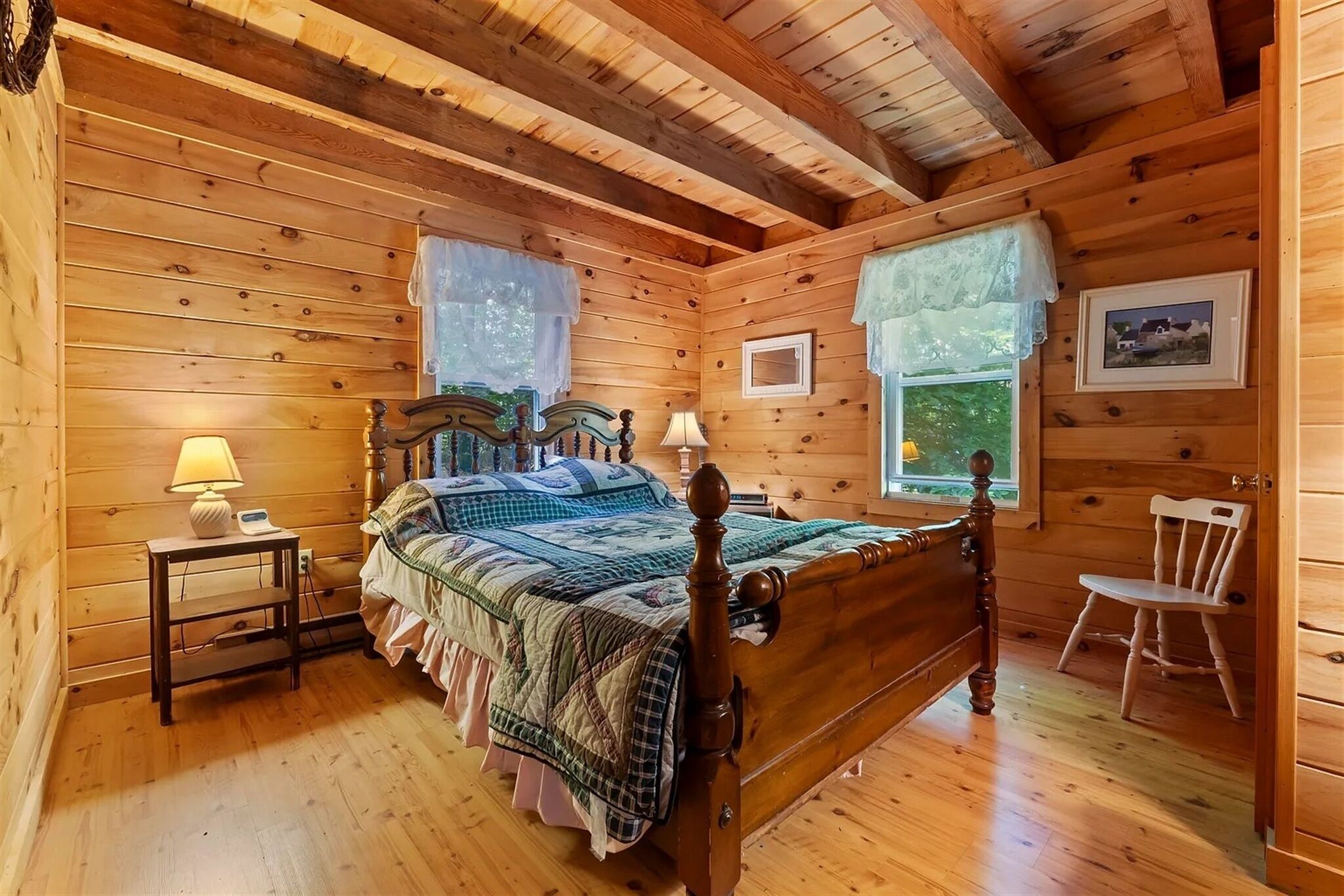 Quarto de cabana de madeira com uma cama de dossel, uma mesa de cabeceira com abajur, uma cadeira e uma janela com cortina.