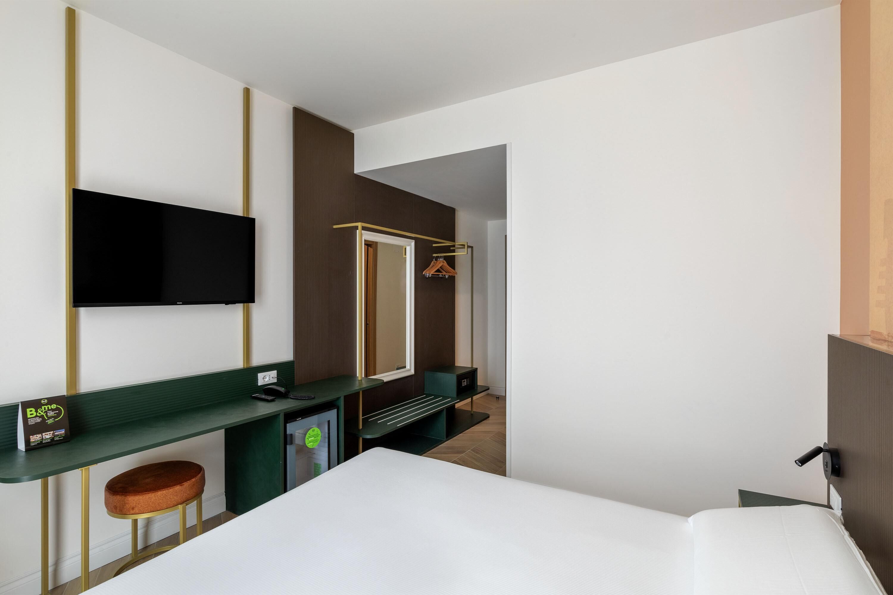 Quarto de hotel com cama, escrivaninha com cadeira, TV de tela plana na parede, espelho e cabideiro.