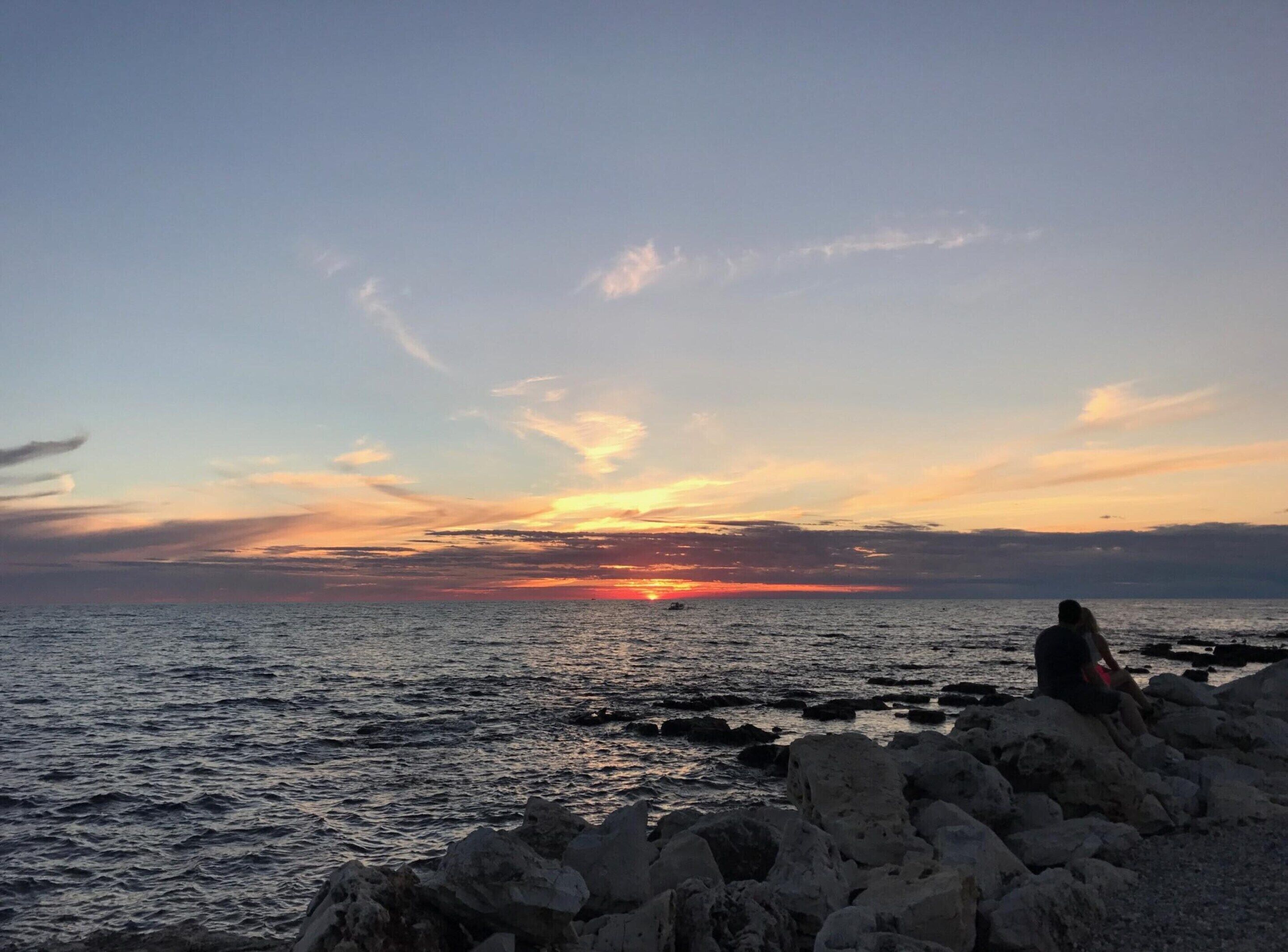 Um pôr do sol sobre uma costa rochosa, com uma pessoa sentada sobre as rochas.