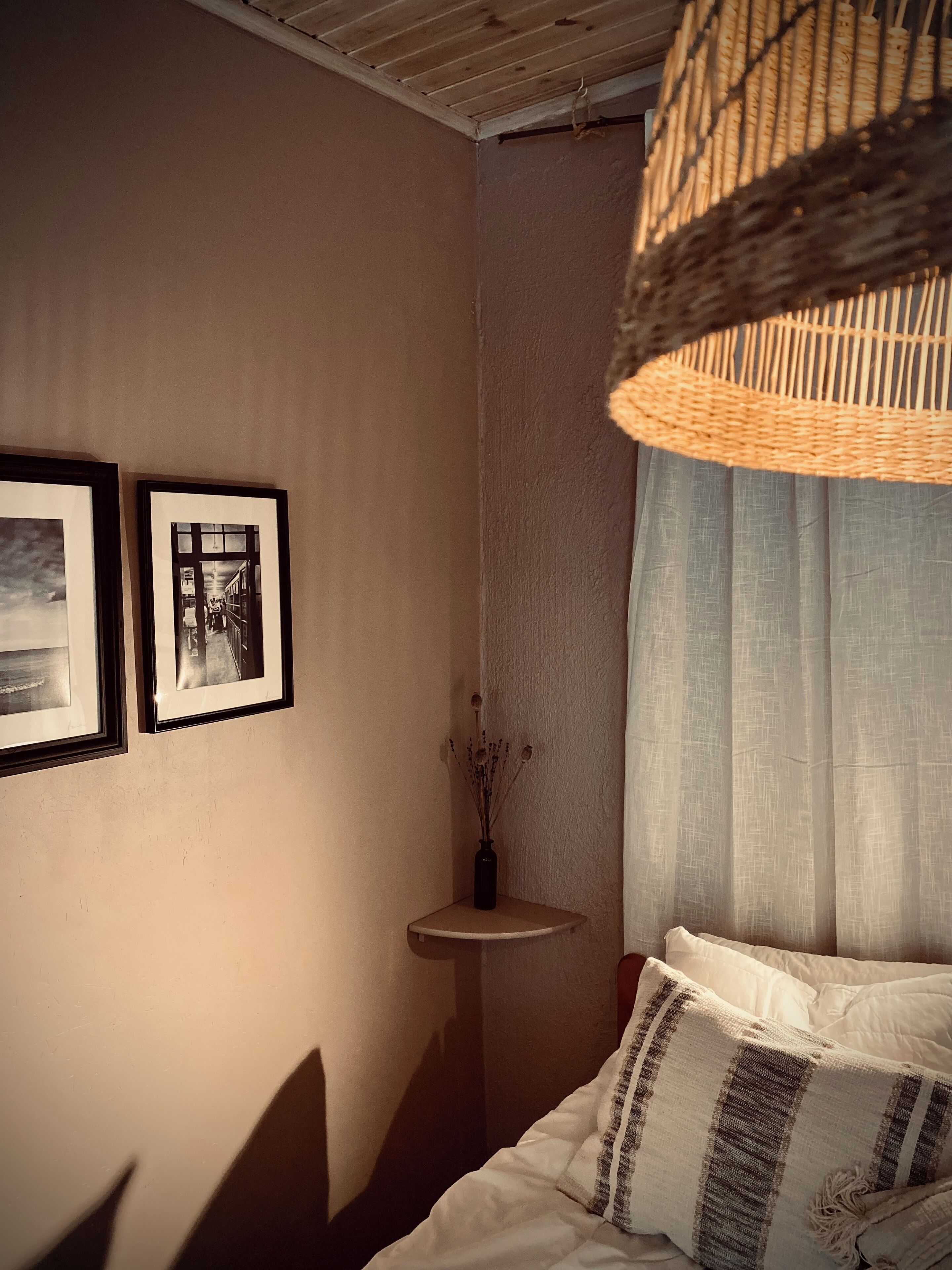 Quarto com cama, mesa de cabeceira pequena com um vaso e duas fotos emolduradas na parede.