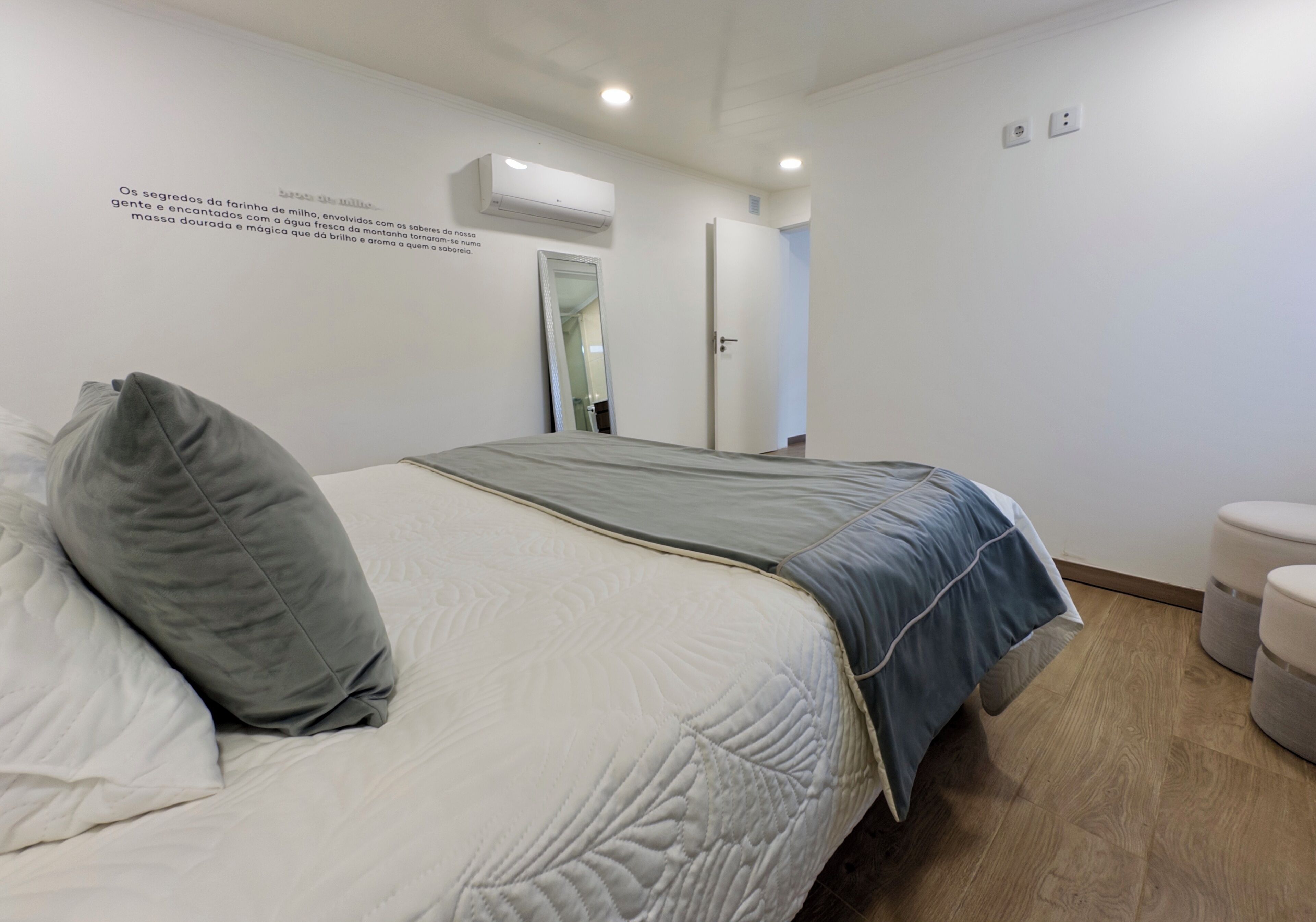Quarto com cama, almofadas, mesa de cabeceira, espelho e porta para outro cômodo.