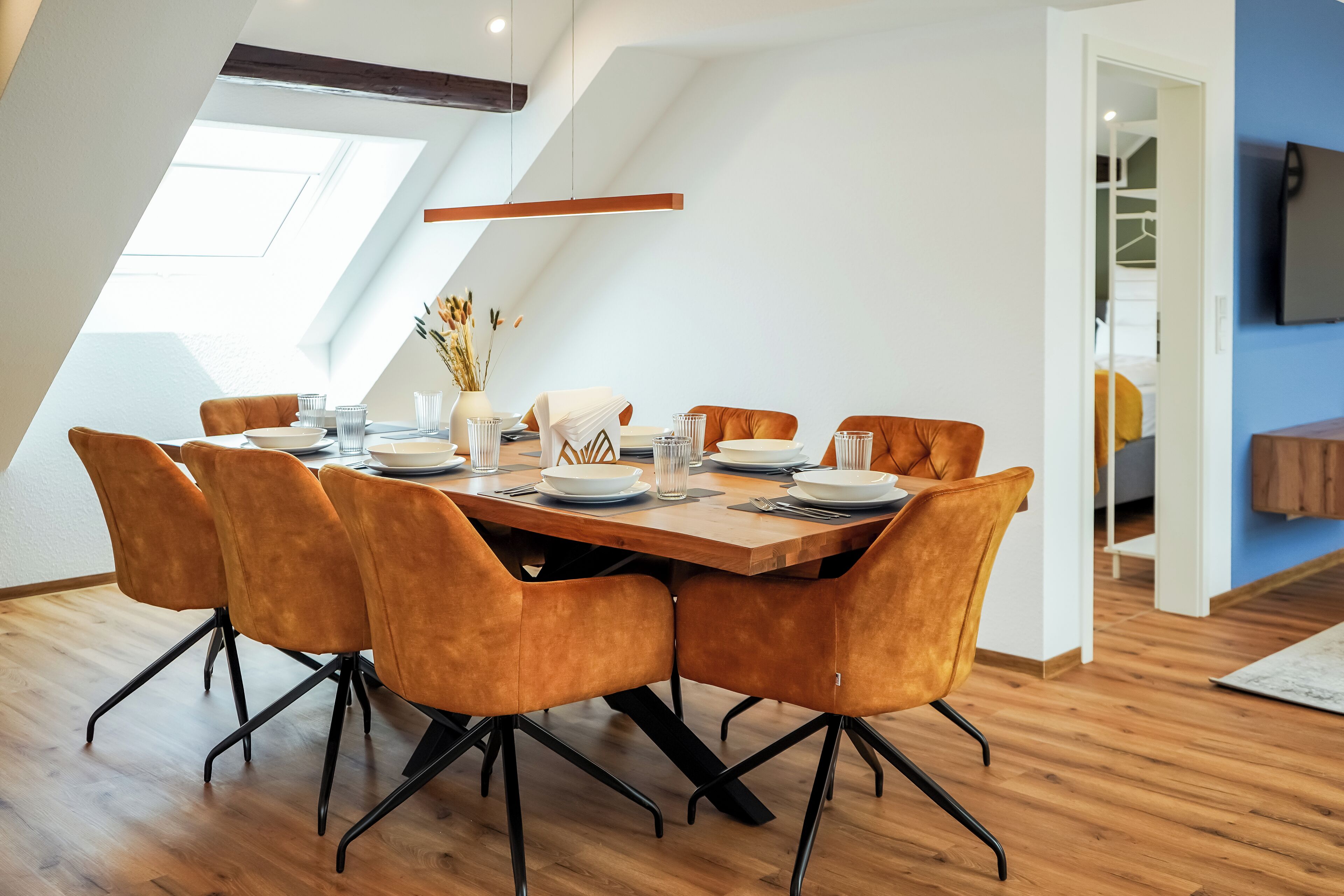 Uma sala de jantar com mesa de madeira posta para seis pessoas, rodeada por seis cadeiras laranjas com pés pretos.