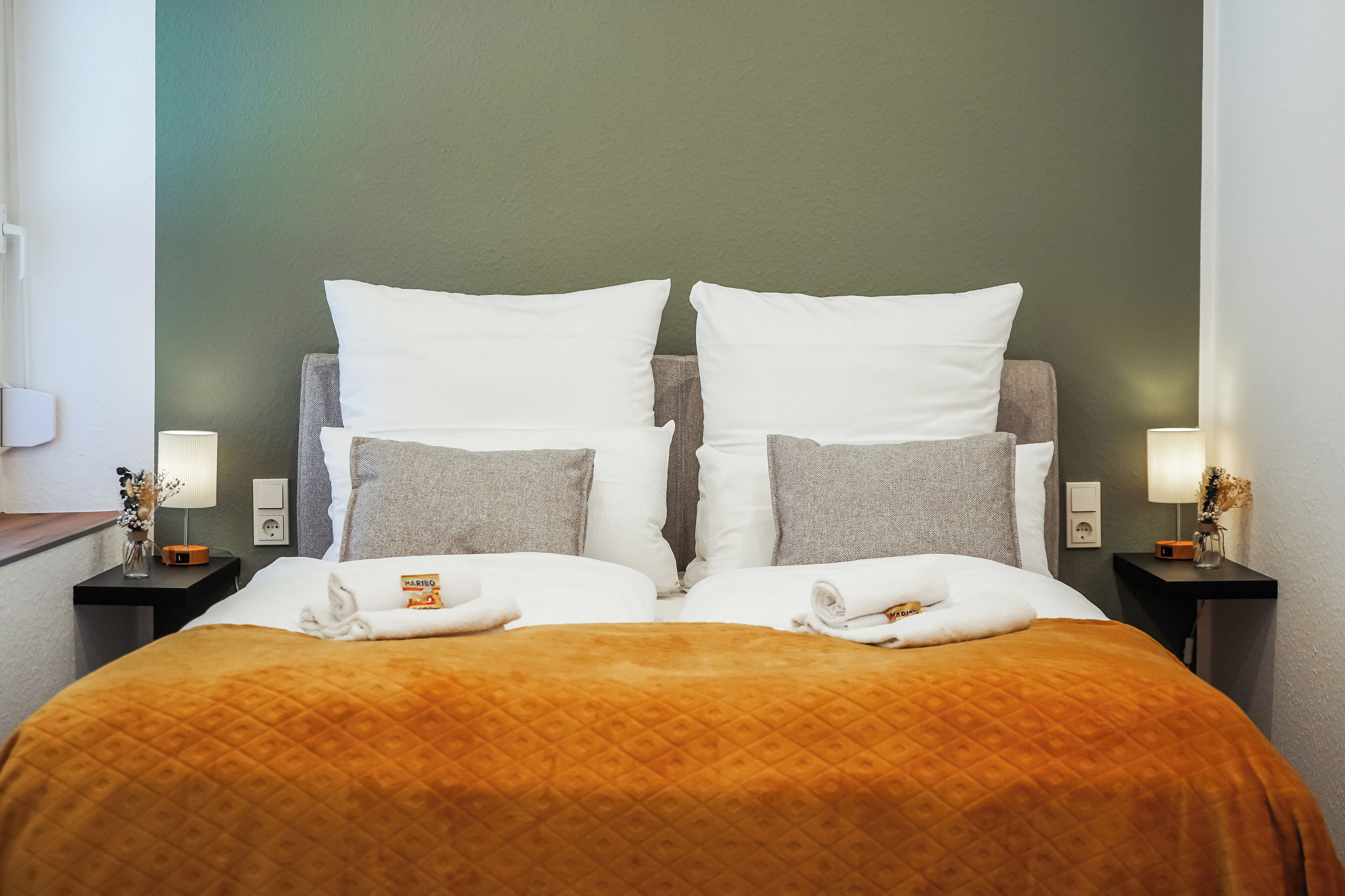 Cama de casal com um cobertor laranja acolchoado, duas almofadas brancas e uma almofada combinando de cada lado. Há duas mesas de cabeceira com luminárias e um pequeno vaso de flores.