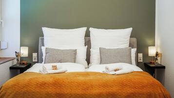 Cama de casal com um cobertor laranja acolchoado, duas almofadas brancas e uma almofada combinando de cada lado. Há duas mesas de cabeceira com luminárias e um pequeno vaso de flores.