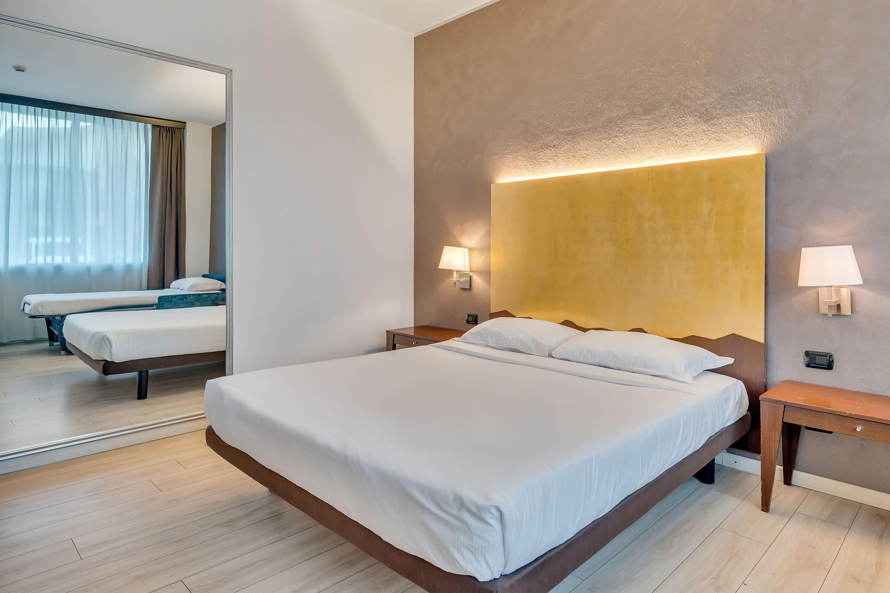 Quarto de hotel com uma cama grande, mesas de cabeceira, um espelho e uma luminária na parede.