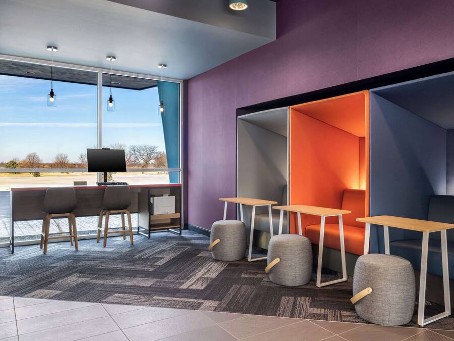 Um espaço de escritório moderno com área de mesa, dois bancos cinzas, uma mesinha e um espaço de estar acolchoado com uma almofada colorida.