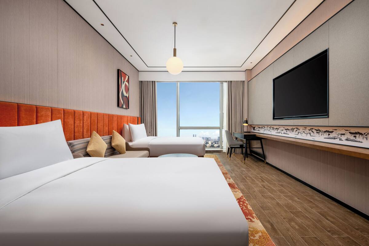 Quarto de hotel com duas camas, uma televisão grande, vista para a cidade e uma escrivaninha com cadeira.