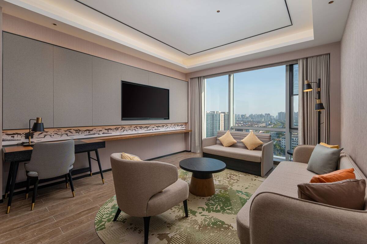 Quarto de hotel moderno com televisão de tela plana na parede, um sofá, uma escrivaninha com cadeira e uma janela grande com vista para a cidade.