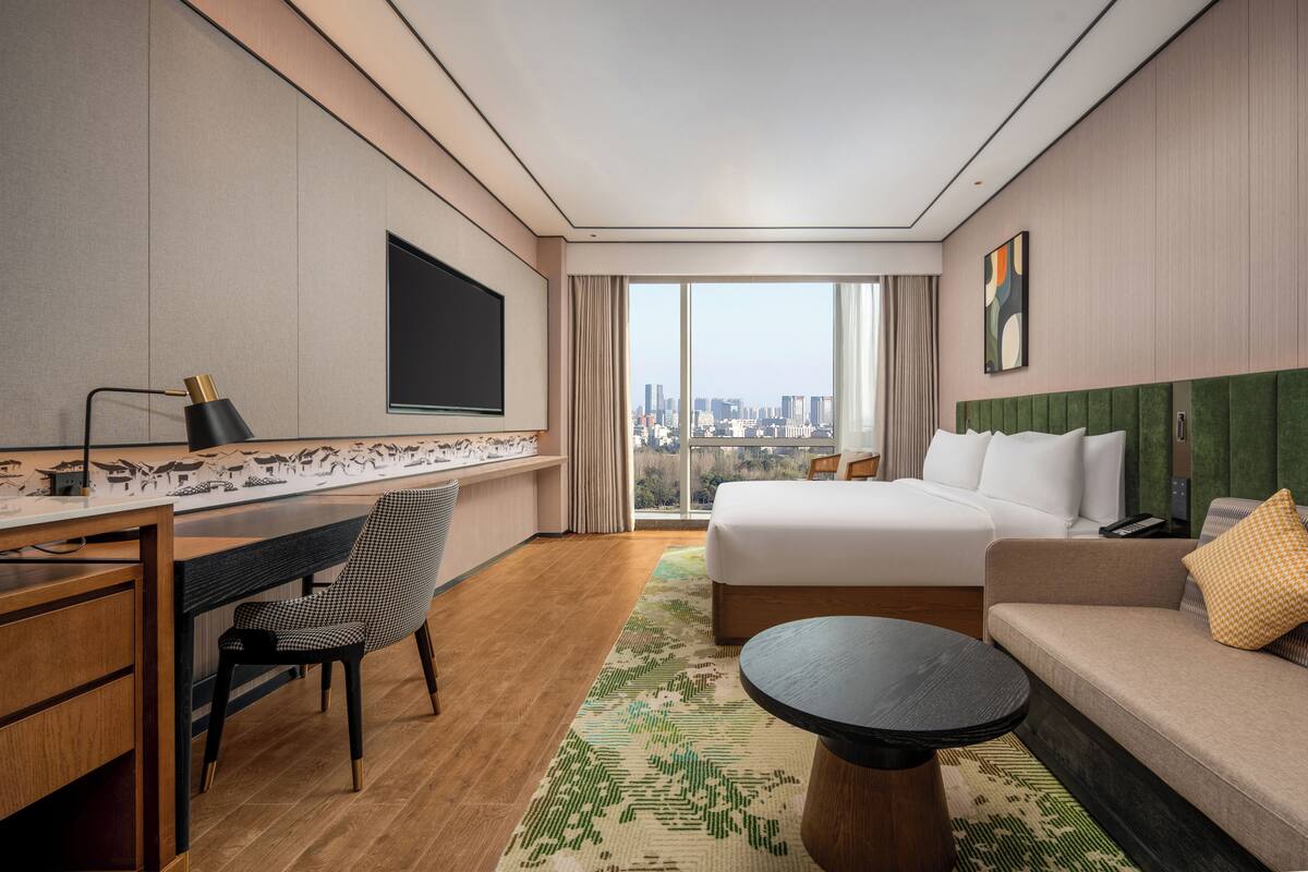 Quarto de hotel com cama, escrivaninha com cadeira, sofá, mesa redonda e uma grande janela com vista para a cidade.