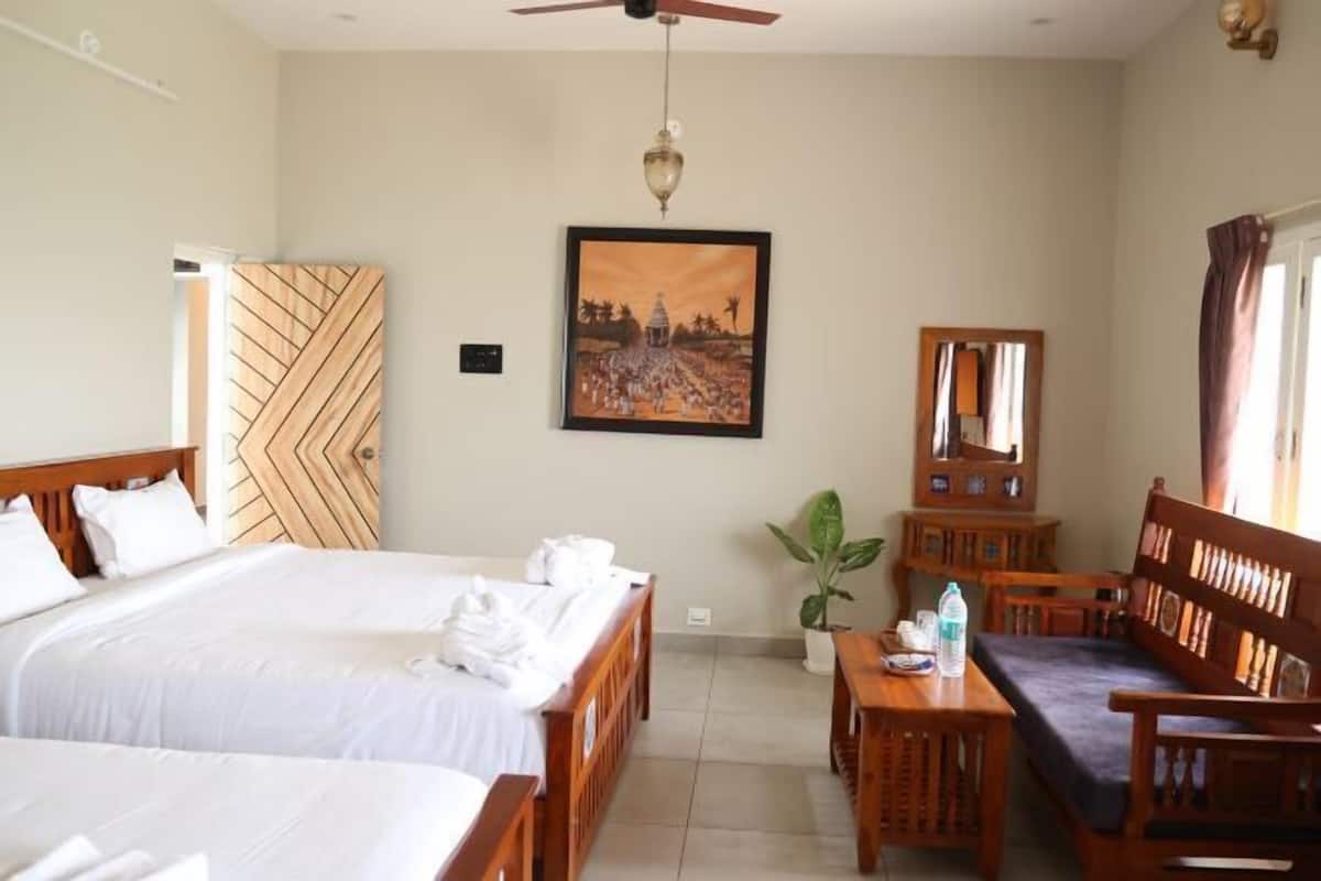 Quarto de hotel com duas camas, um ventilador de teto, um quadro emoldurado na parede, um vaso de plantas e um espaço de estar com móveis de madeira.