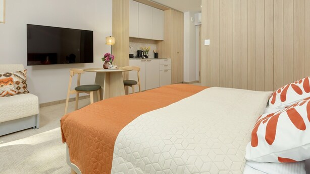 Quarto de hotel com cama, sofá, televisão, pequena mesa de jantar e cozinha compacta.