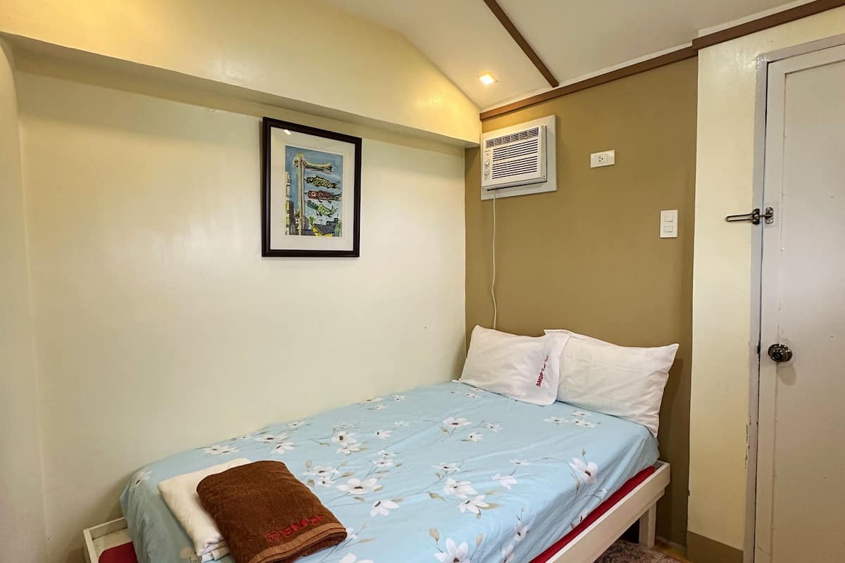 Uma cama de solteiro com colcha estampada, uma toalha marrom, um quadro na parede e um aparelho de ar condicionado.
