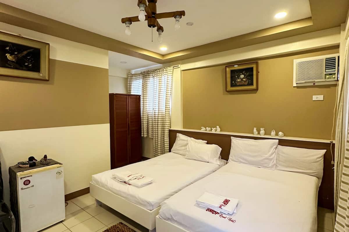 Quarto de hotel com duas camas, um pequeno frigorífico, um guarda-roupa de madeira e um candeeiro de teto.