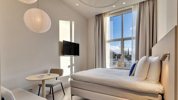 Quarto de hotel moderno com uma cama grande, uma televisão, uma pequena mesa e vista para a cidade.