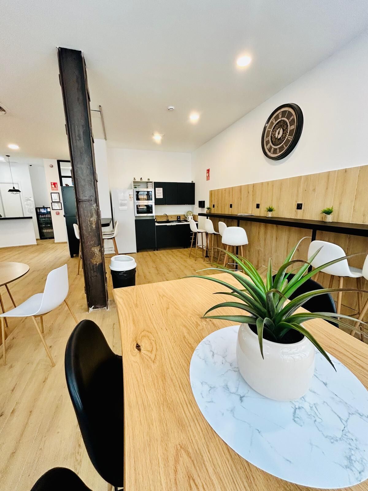 Um interior moderno com mesa de jantar de madeira, cadeiras brancas, uma planta em vaso e uma área de cozinha ao fundo.