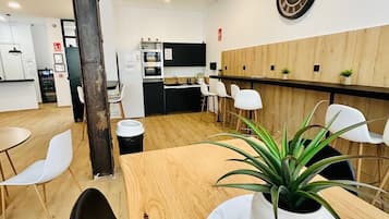 Um interior moderno com mesa de jantar de madeira, cadeiras brancas, uma planta em vaso e uma área de cozinha ao fundo.