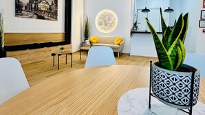 Um espaço de escritório moderno com uma mesa de conferência de madeira, um sofá com almofadas amarelas, uma planta em vaso e uma pintura da paisagem urbana na parede.