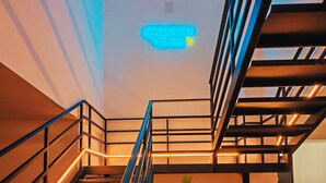 Uma escadaria moderna com guarda-corpos de metal e degraus de madeira, um letreiro de neon com a inscrição "URBAN INN MADRID" e uma planta em vaso no centro.
