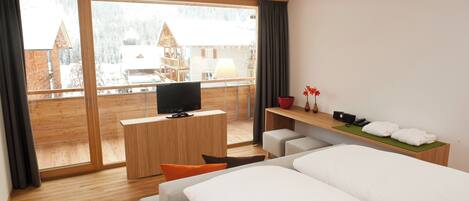 Quarto de hotel com cama, escrivaninha, televisão e varanda com vista para edifícios e árvores.