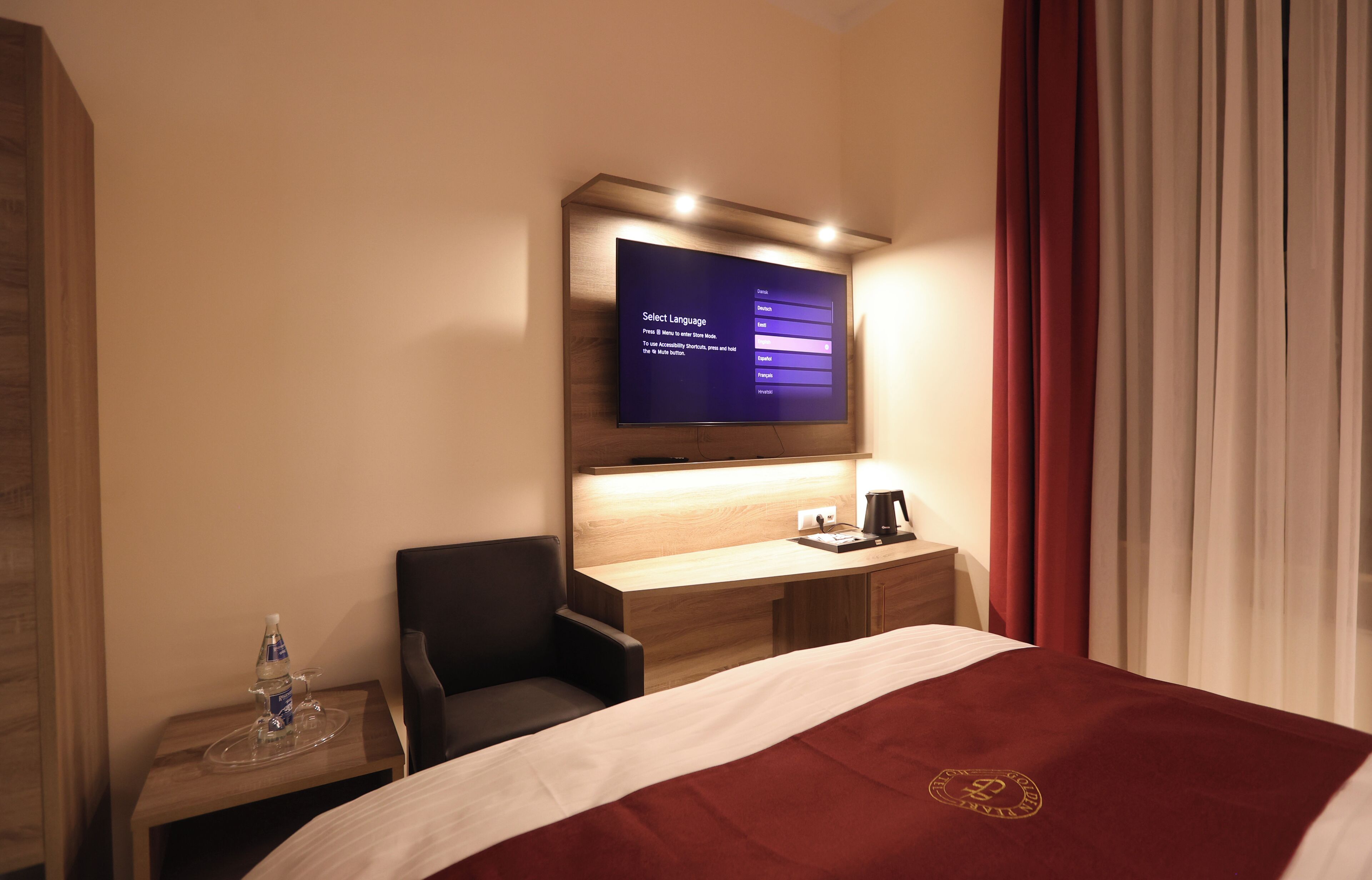 Quarto de hotel com cama, escrivaninha, cadeira, televisão fixada na parede e janela com cortinas.