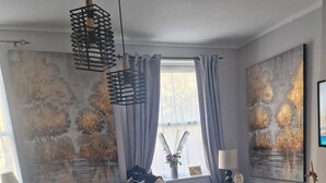 Uma sala de estar com uma mesa de centro branca, um sofá azul, uma poltrona azul e um banquinho azul. Há um quadro na parede, um abajur sobre uma mesinha lateral e uma planta no parapeito da janela.