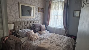 Quarto com cabeceira estofada, cama grande com várias almofadas, mala e janela com cortinas finas.