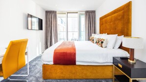 Quarto de hotel com uma cama grande, escrivaninha, cadeira, televisão e janela com cortinas.