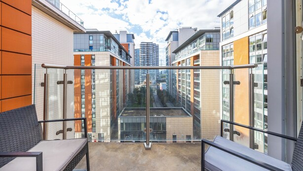 Uma varanda com guarda-corpo de vidro, um banco acolchoado e vista para edifícios modernos e um céu claro.