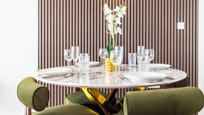 Mesa redonda para refeições com pratos brancos, copos de vidro transparente e um arranjo de flores brancas. A mesa está posicionada em frente a uma parede com ripas de madeira verticais e uma luminária moderna.
