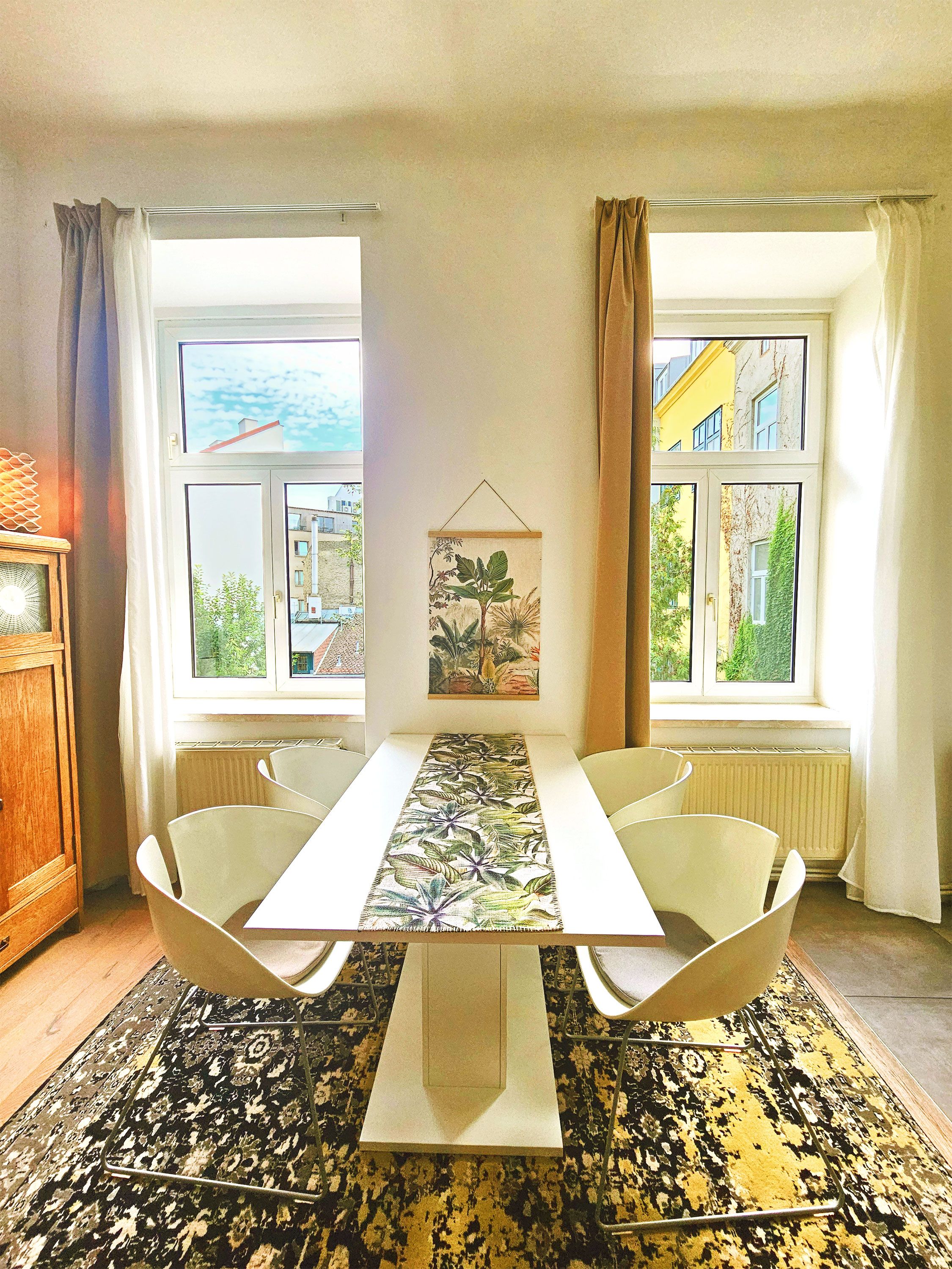 Uma área de refeições com mesa e cadeiras, uma impressão botânica pendurada e vista para edifícios através de grandes janelas.