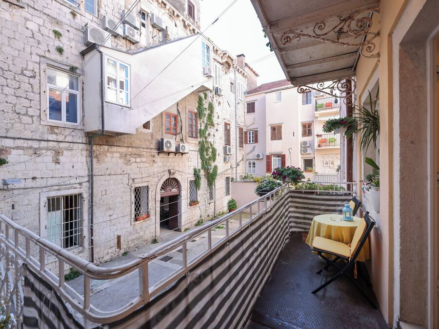 Uma viela estreita com uma pequena área de estar ao ar livre em uma varanda, cercada por edifícios antigos com varandas e janelas.