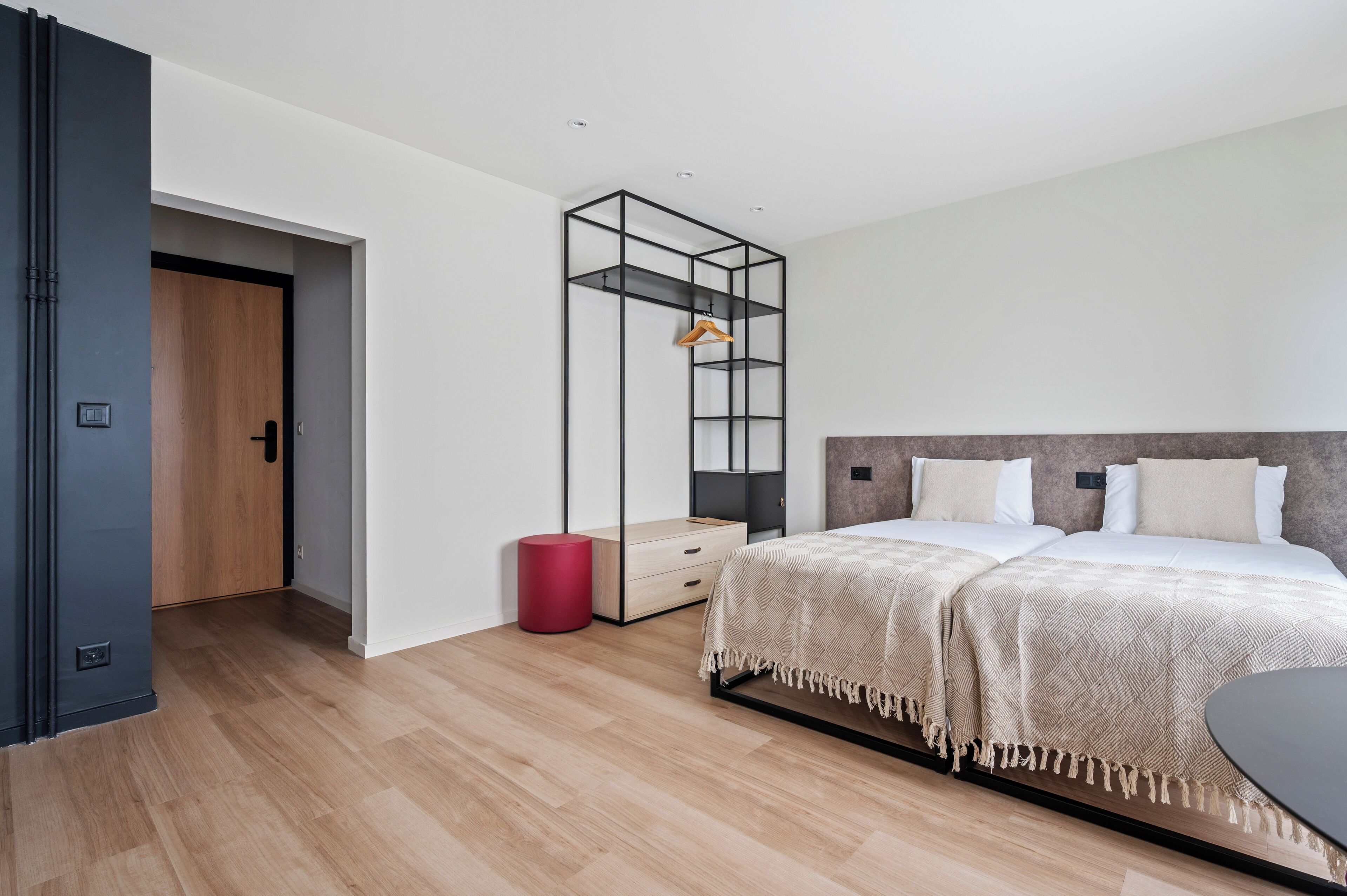 Quarto moderno com piso de madeira, cama com cobertor bege, lixeira vermelha e estante de metal preta.