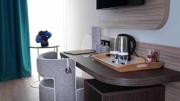 Quarto de hotel pequeno com televisão de tela plana na parede, uma mesinha com bandeja contendo copos e chaleira, uma cadeira cinza e uma lixeira.