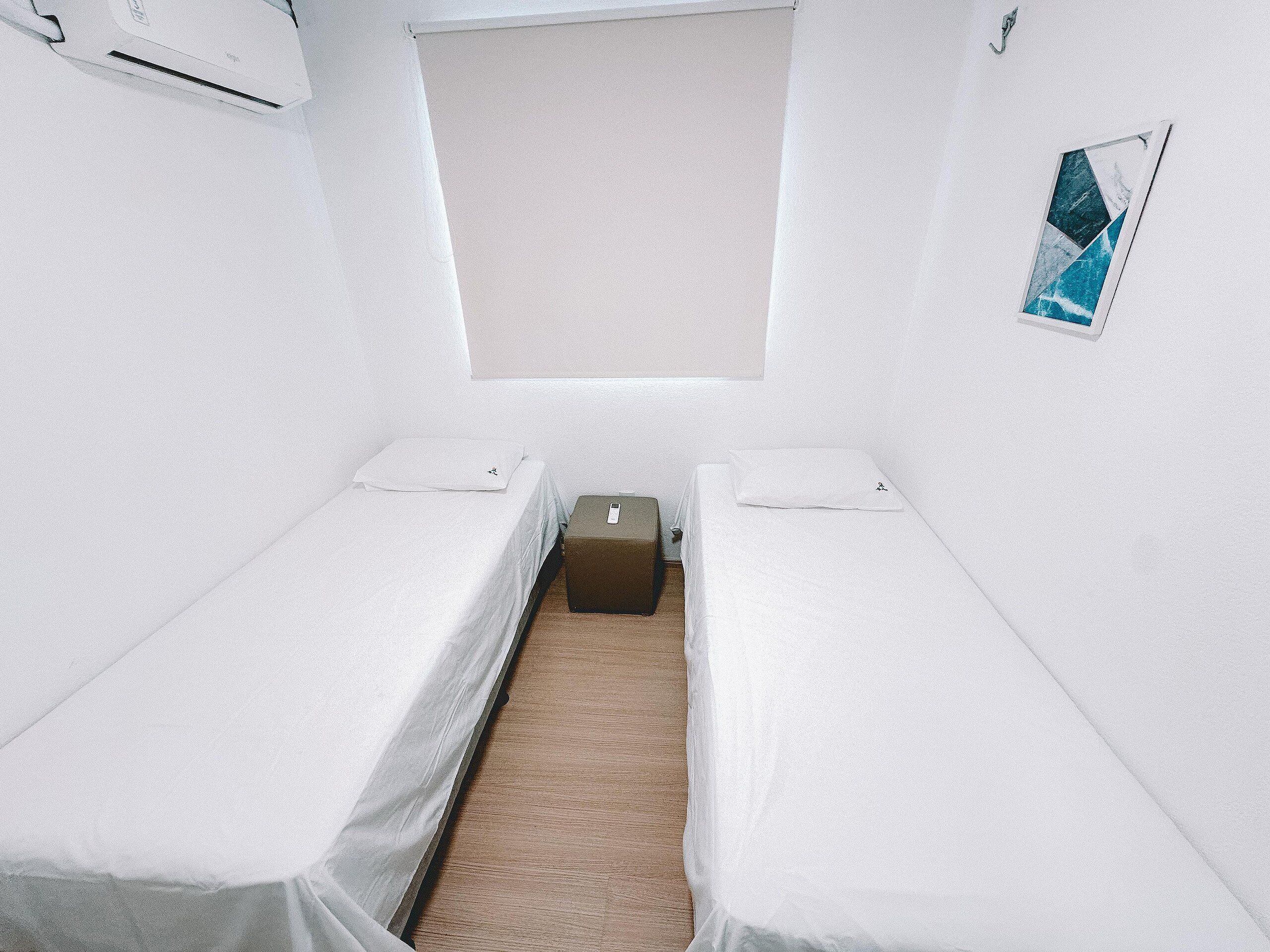 Quarto pequeno e limpo, com duas camas de solteiro, uma janela e ar condicionado fixado na parede.