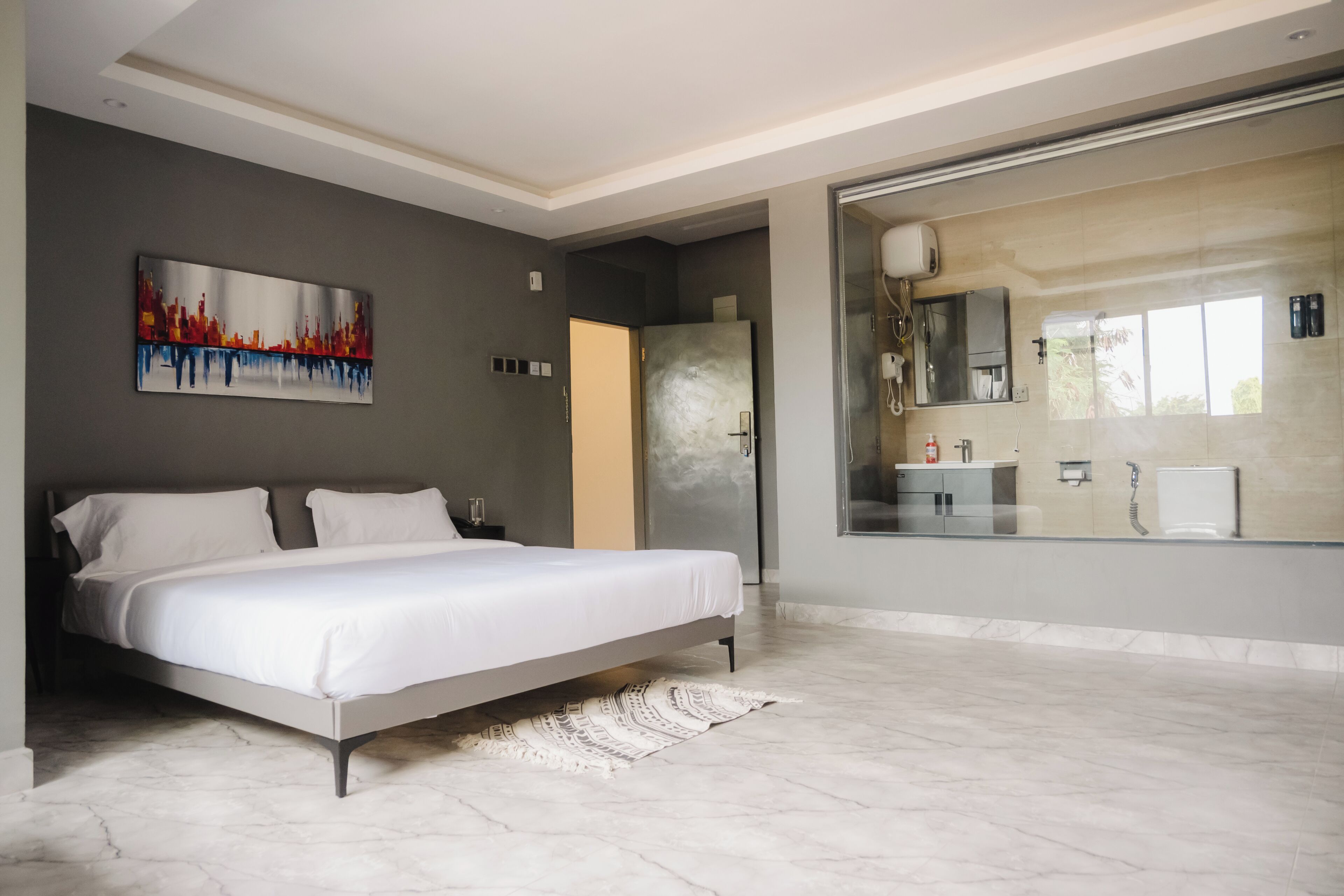 Quarto moderno com cama grande, pintura de paisagem urbana, banheiro com chuveiro de vidro e piso de mármore.